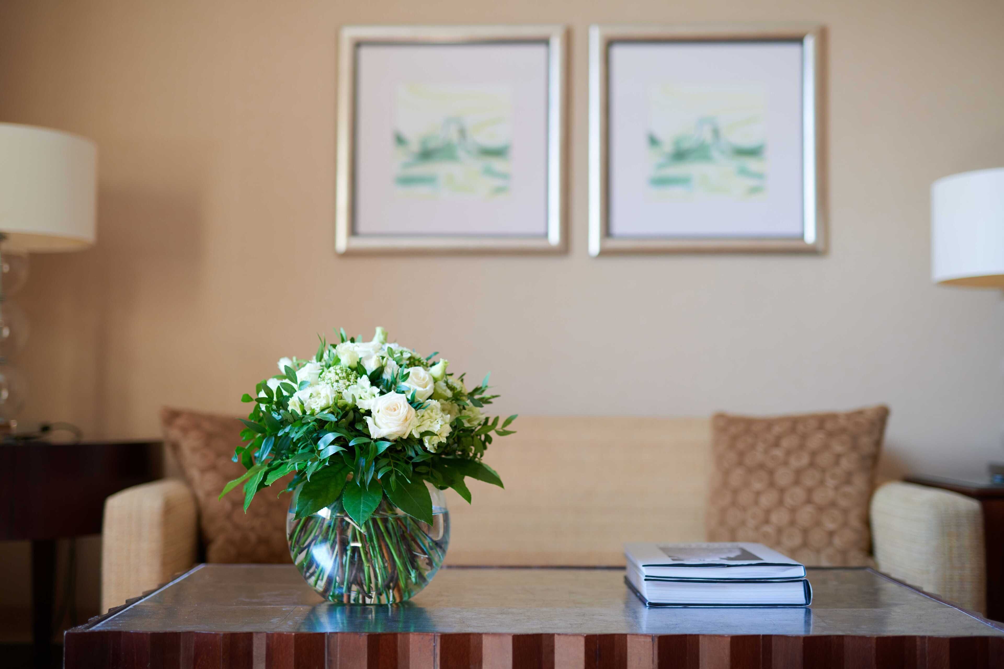 Um quarto com uma mesa de centro de madeira, um vaso com flores brancas, um sofá com almofadas estampadas e duas fotos emolduradas na parede.