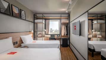 Quarto de hotel com duas camas, uma escrivaninha, uma cadeira, uma mesinha, um espelho e uma janela com vista.