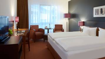 Quarto de hotel com uma cama grande, duas poltronas, uma escrivaninha com um laptop, uma televisão e uma pequena mesa com um objeto decorativo.