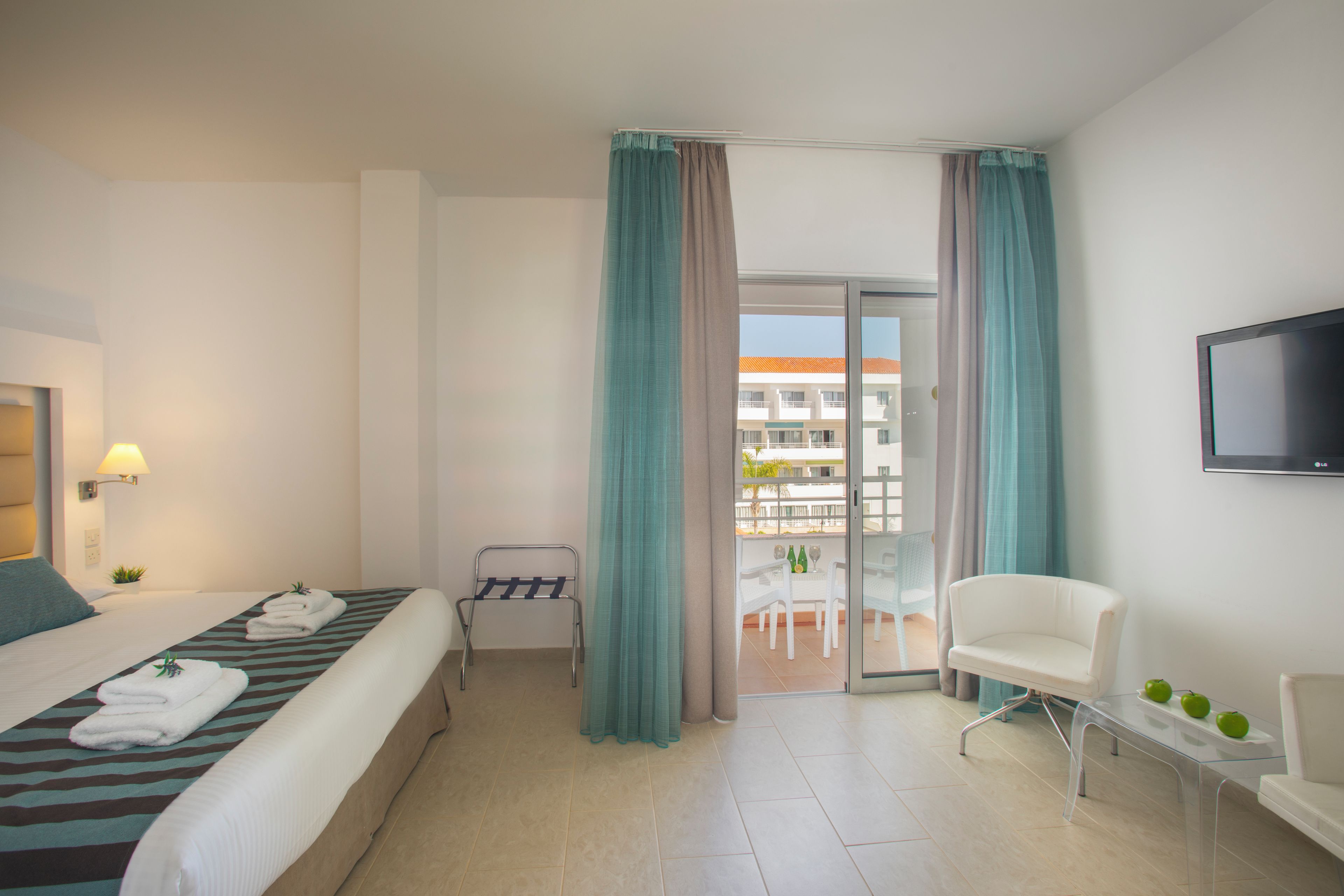 Quarto de hotel com cama, área de estar com mesa e cadeiras, televisão na parede e varanda com cortinas.
