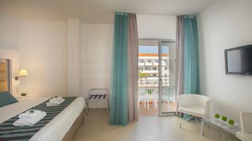 Quarto de hotel com cama, área de estar com mesa e cadeiras, televisão na parede e varanda com cortinas.