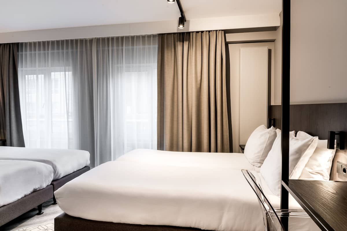 Quarto de hotel com duas camas, uma janela grande com cortinas leves e uma mesa de cabeceira com abajur.