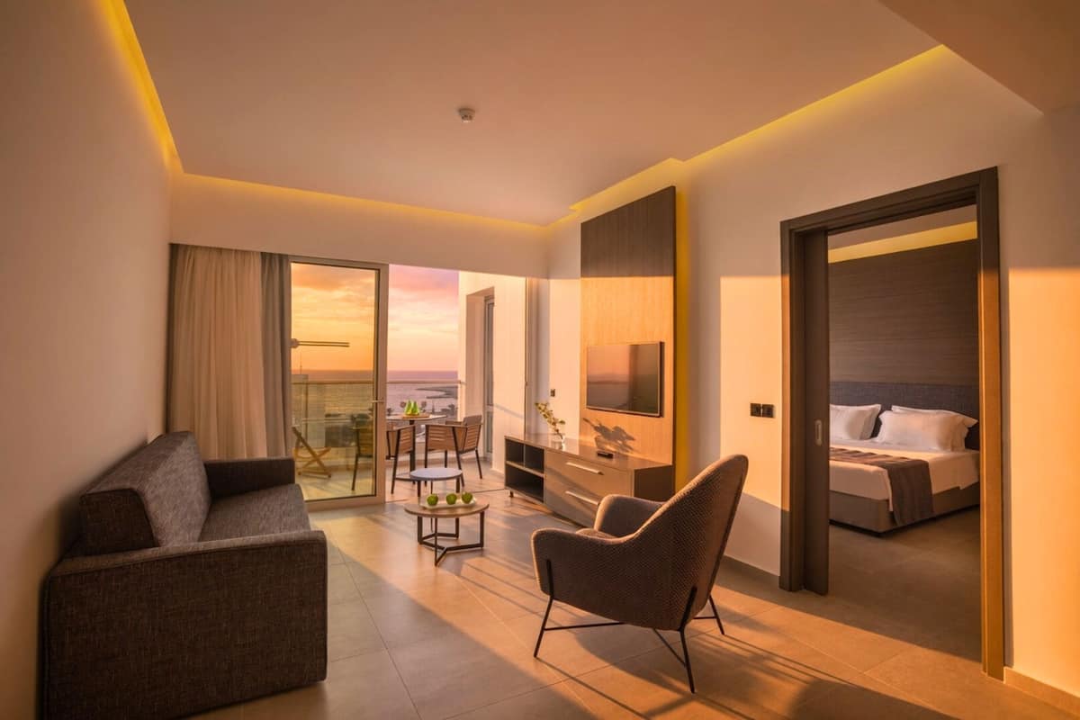 Quarto de hotel moderno com varanda, sofá, poltrona, televisão e cama.