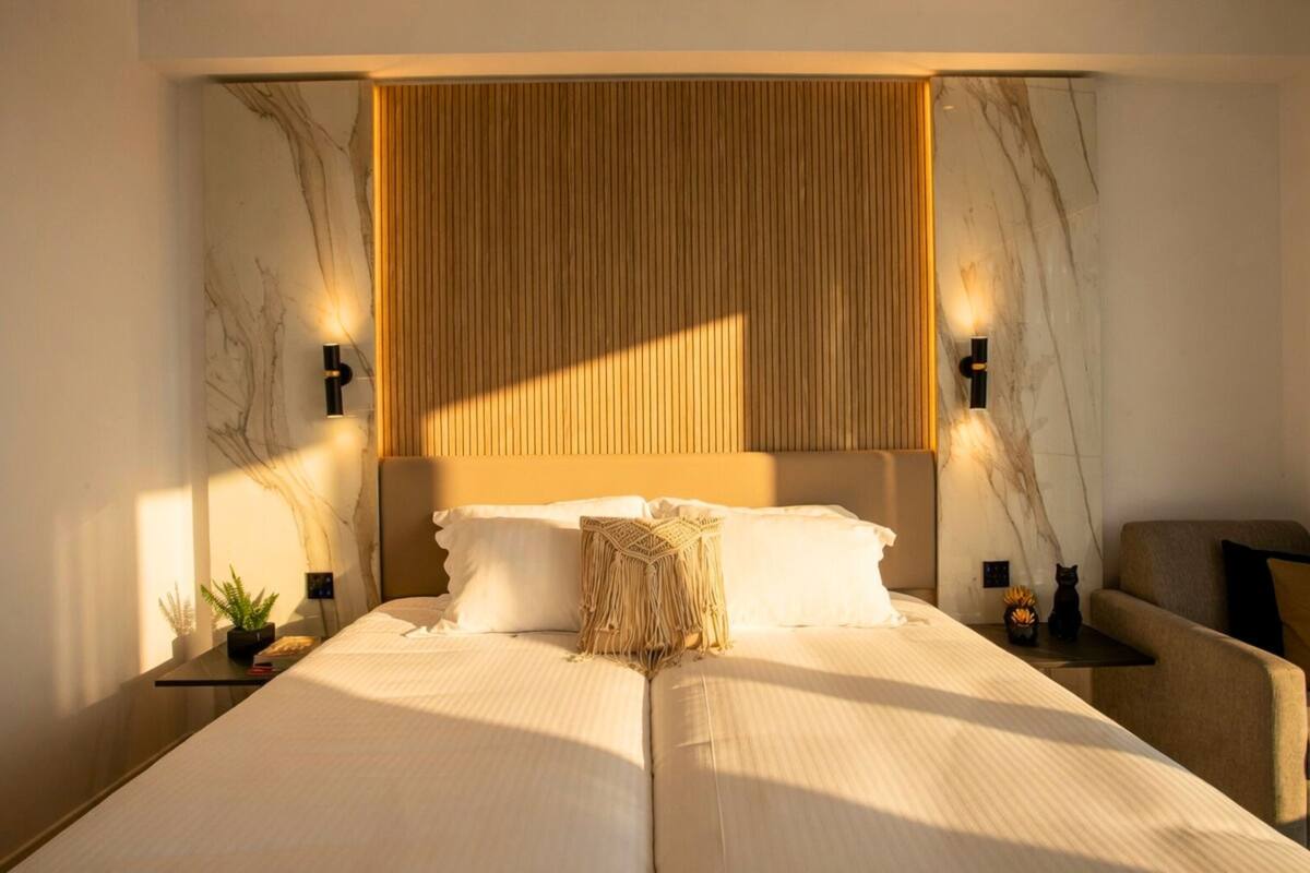 Uma cama de casal com cabeceira de madeira, mesas de cabeceira com abajures e um vaso com planta.