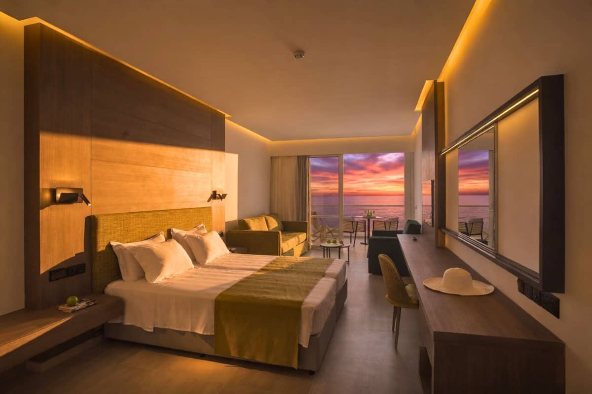 Quarto de hotel moderno com uma cama grande, uma escrivaninha, uma cadeira e vista para o pôr do sol através de amplas janelas.
