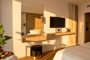 Quarto de hotel moderno com uma cama grande, televisão montada, mesa de cabeceira com planta e um espelho refletindo o interior do quarto.