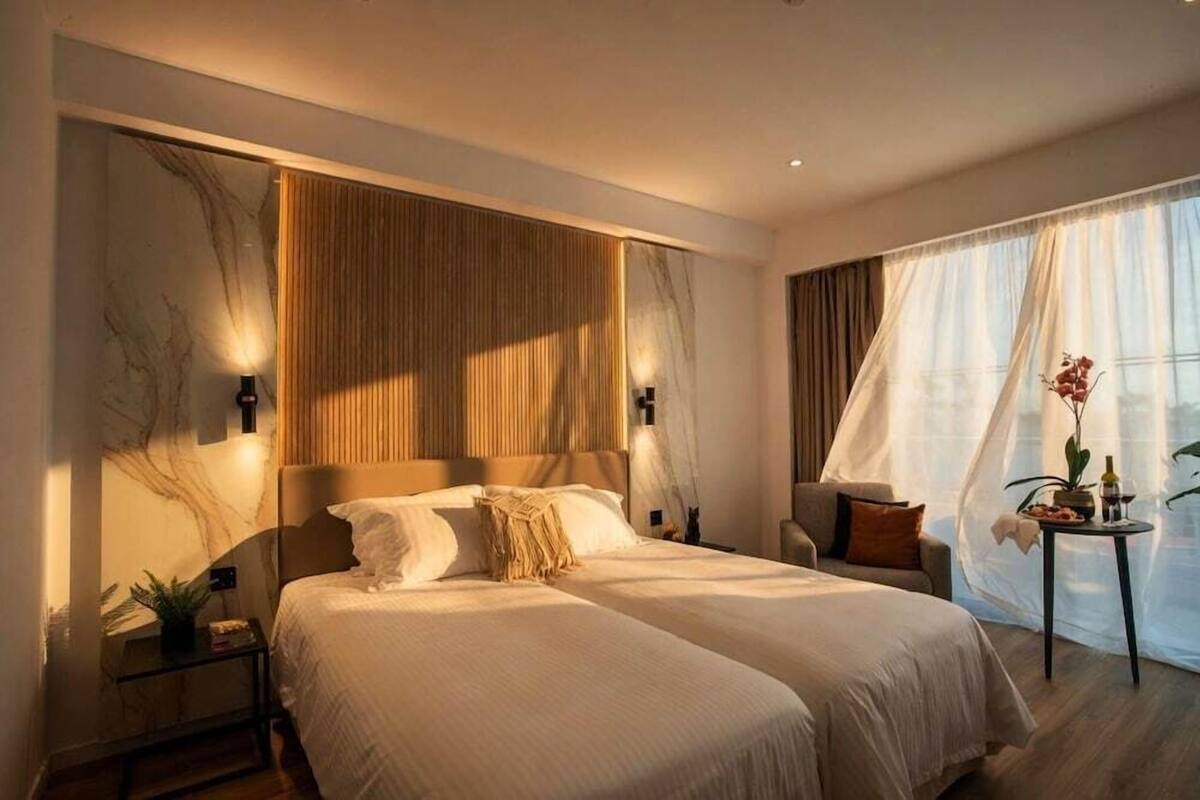 Quarto de hotel moderno com uma cama grande, uma mesa de cabeceira, uma pequena mesa com uma planta e uma janela com cortinas translúcidas.