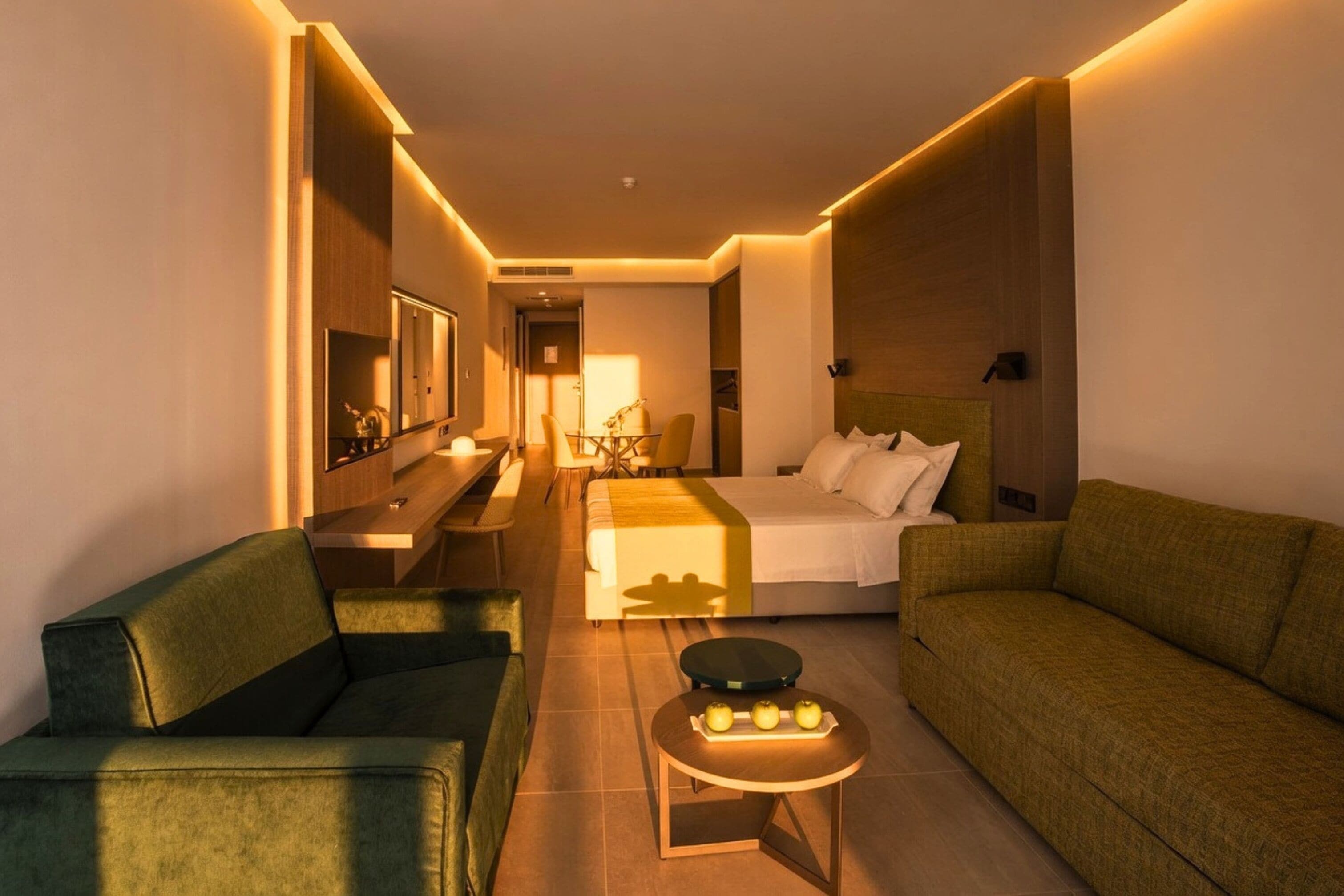 Quarto de hotel com uma cama, dois sofás verdes, uma mesinha com maçãs, uma escrivaninha com abajur e uma televisão.