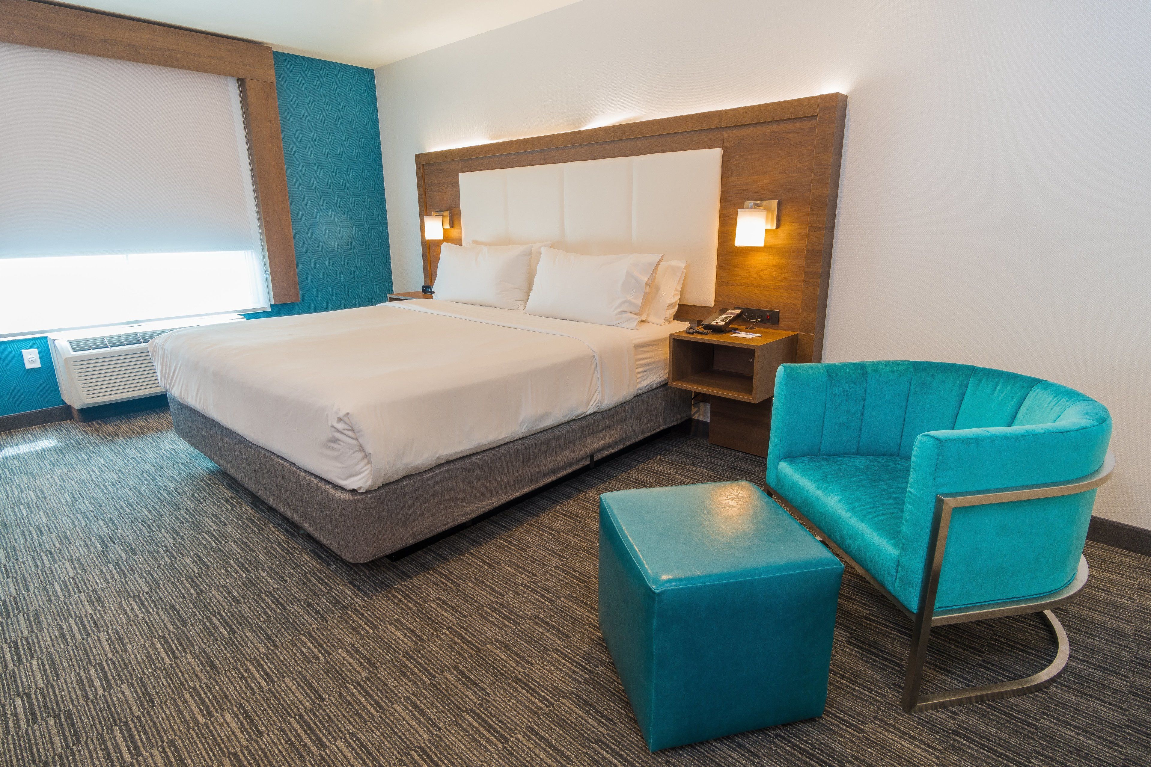 Quarto de hotel com uma cama grande, uma poltrona azul-esverdeada, um pequeno banquinho e uma mesa de cabeceira com telefone, além de uma luminária na parede.