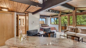 Interior moderno com teto de madeira, cozinha com bancada de mármore, sofá e cama com vista para o exterior.