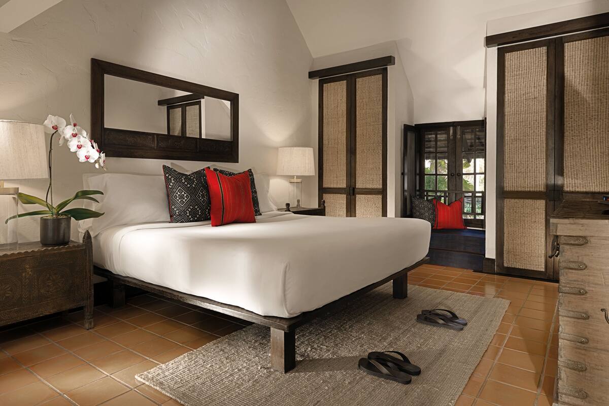 Quarto com cama grande, mesa de cabeceira com abajur, planta em vaso e vista para o exterior através de uma porta.