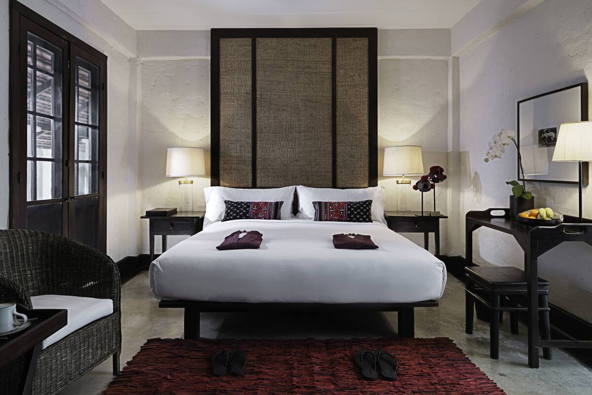 Quarto de hotel com uma cama grande, duas mesas de cabeceira com abajures, uma cadeira, uma mesinha com um vaso e frutas, e um tapete vermelho.
