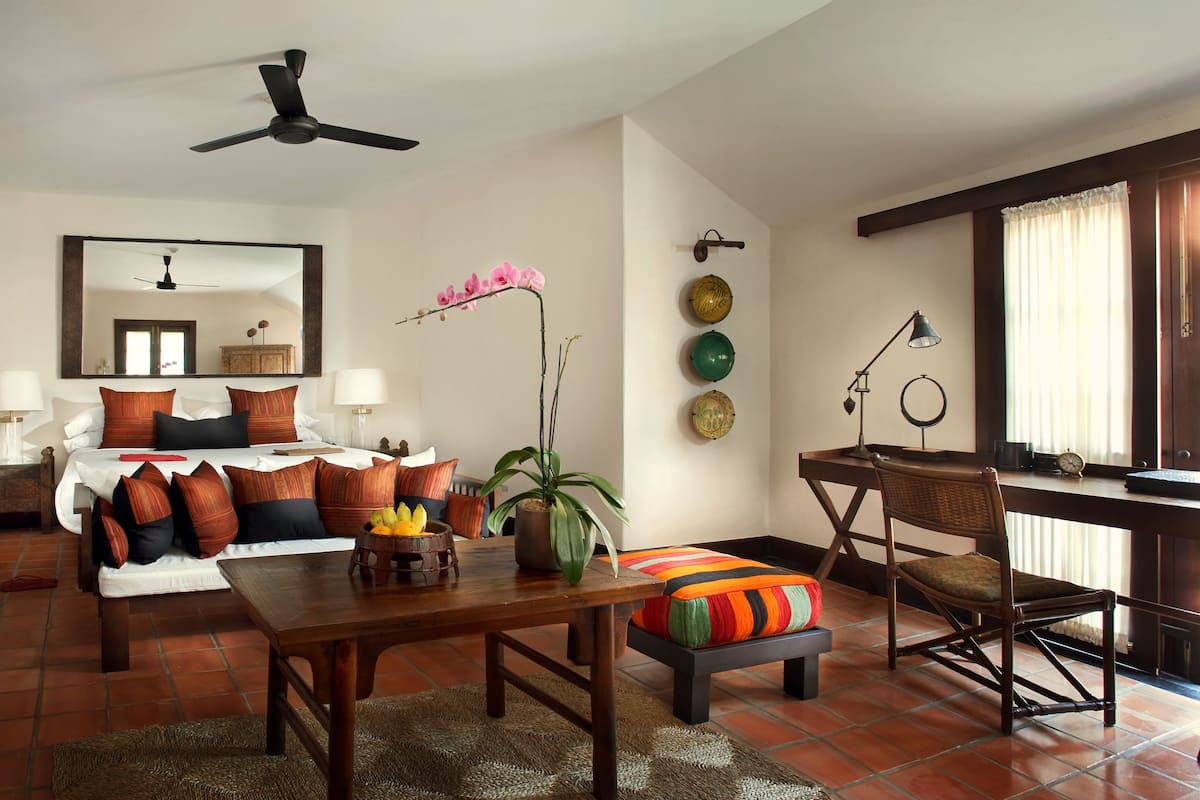 Quarto de hotel com cama, escrivaninha, cadeira, mesa de jantar, planta em vaso e ventoinha de teto.