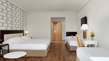 Quarto de hotel com duas camas, uma mesinha, uma cadeira, uma escrivaninha com abajur e televisão na parede.