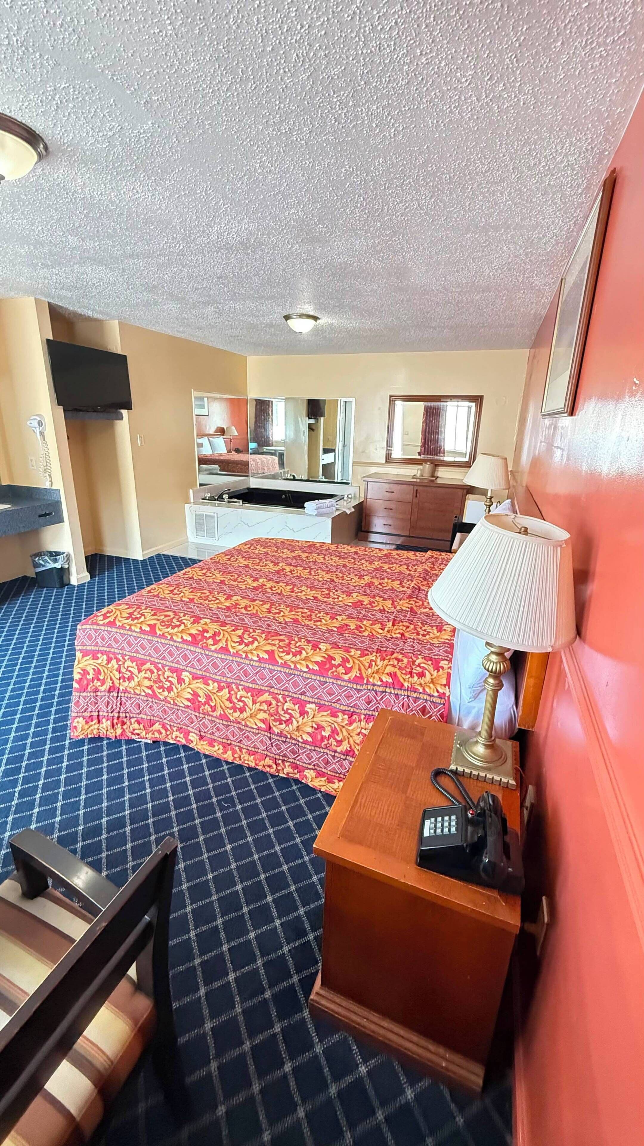 Quarto de hotel com cama, escrivaninha com luminária, televisão e banheiro visível por uma porta aberta.