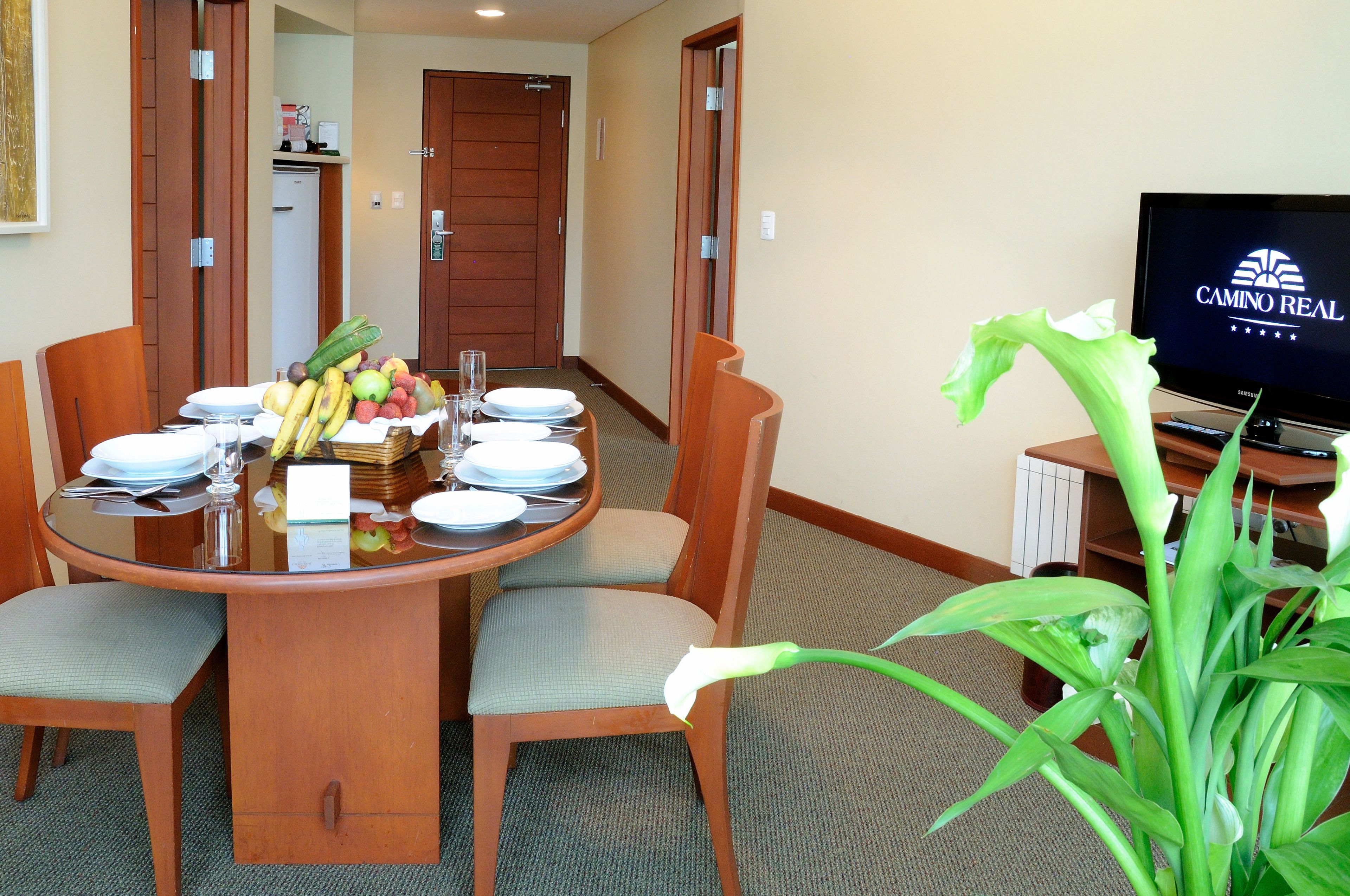 Um espaço de refeições de hotel com uma mesa redonda posta para quatro pessoas, uma televisão exibindo 'Camino Real' e uma grande planta verde.