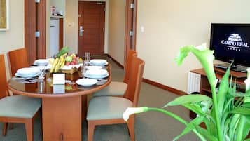 Um espaço de refeições de hotel com uma mesa redonda posta para quatro pessoas, uma televisão exibindo 'Camino Real' e uma grande planta verde.
