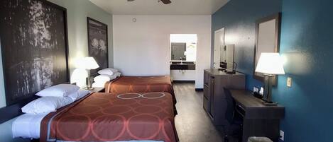 Quarto de hotel com duas camas, um ventilador de teto, uma mesa com luminária, um espelho e vista para o banheiro.