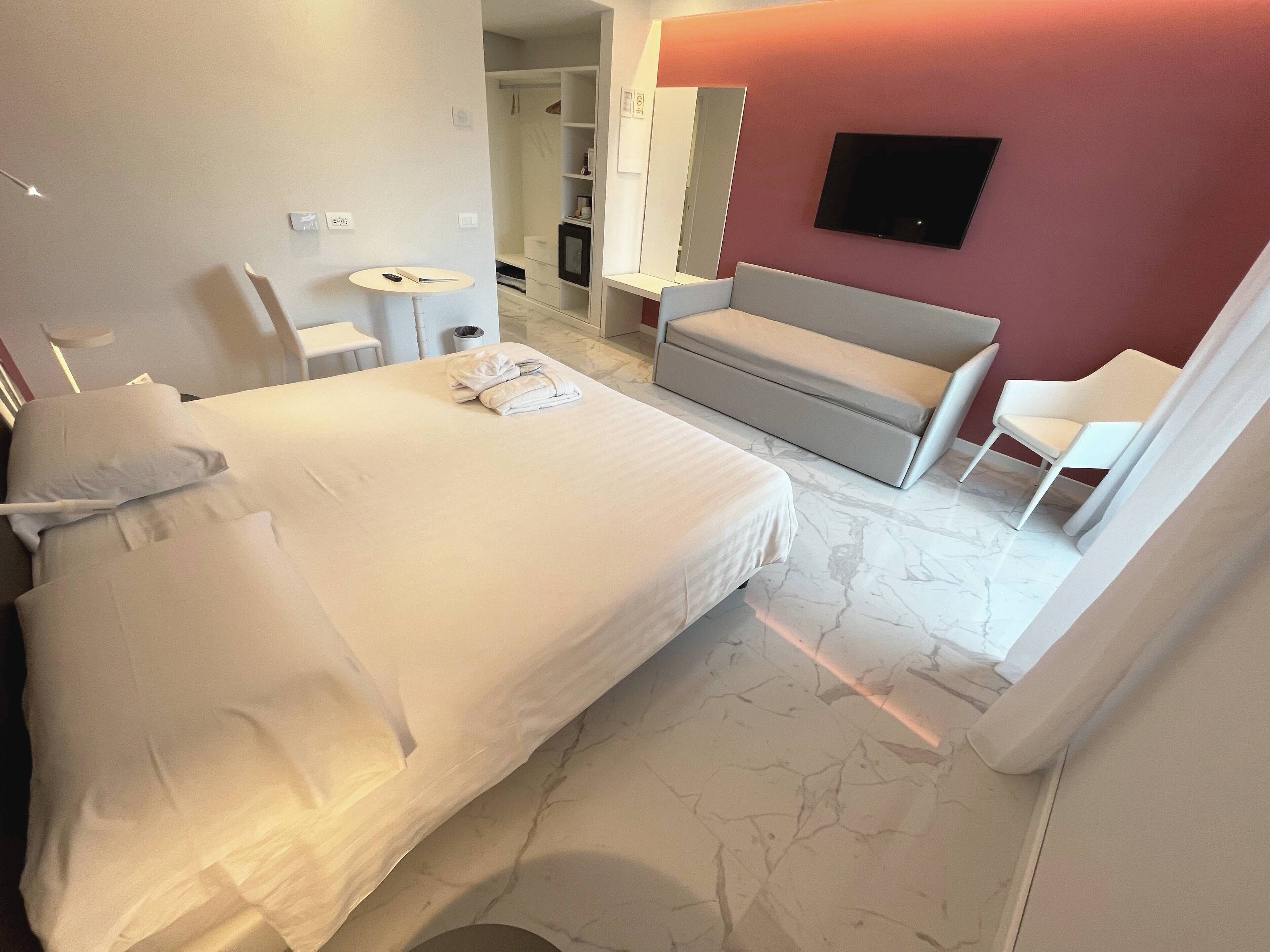 Quarto de hotel com cama grande, sofá, televisão fixada na parede, mesinha e cadeira.
