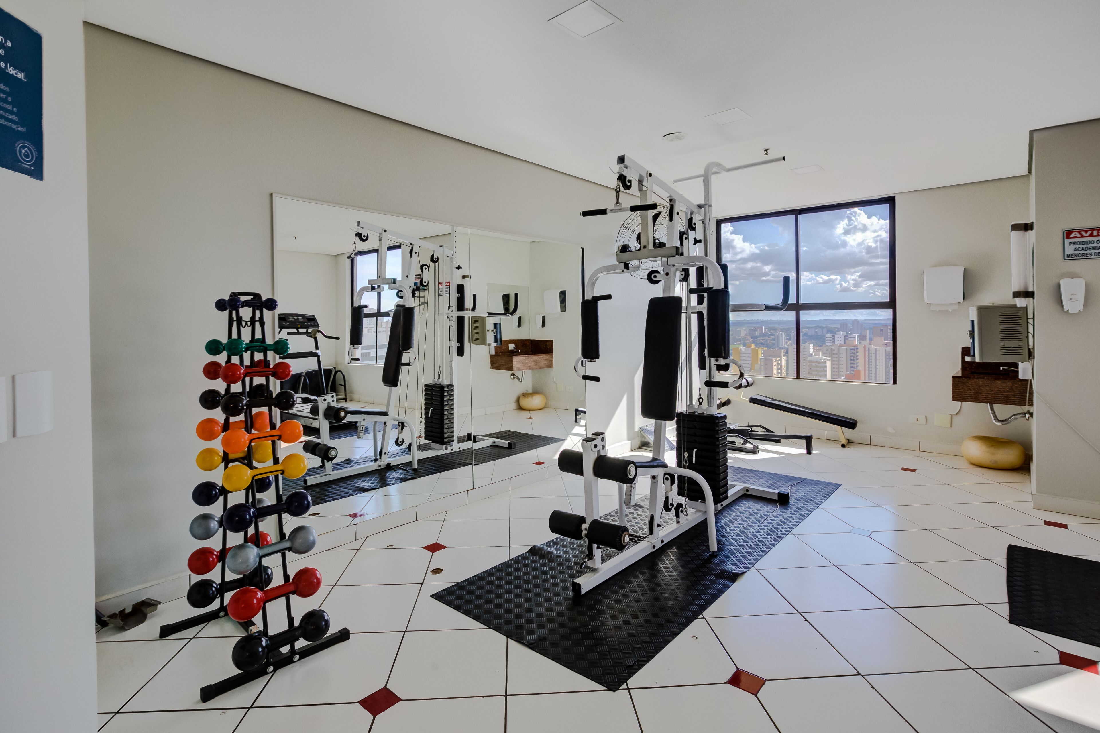 Uma academia com diversos equipamentos de exercício, uma grande janela com vista para a cidade e um tapete preto no chão.