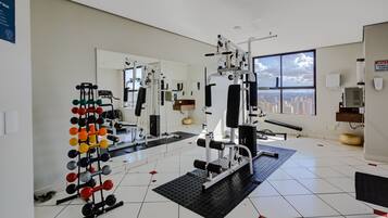 Uma academia com diversos equipamentos de exercício, uma grande janela com vista para a cidade e um tapete preto no chão.