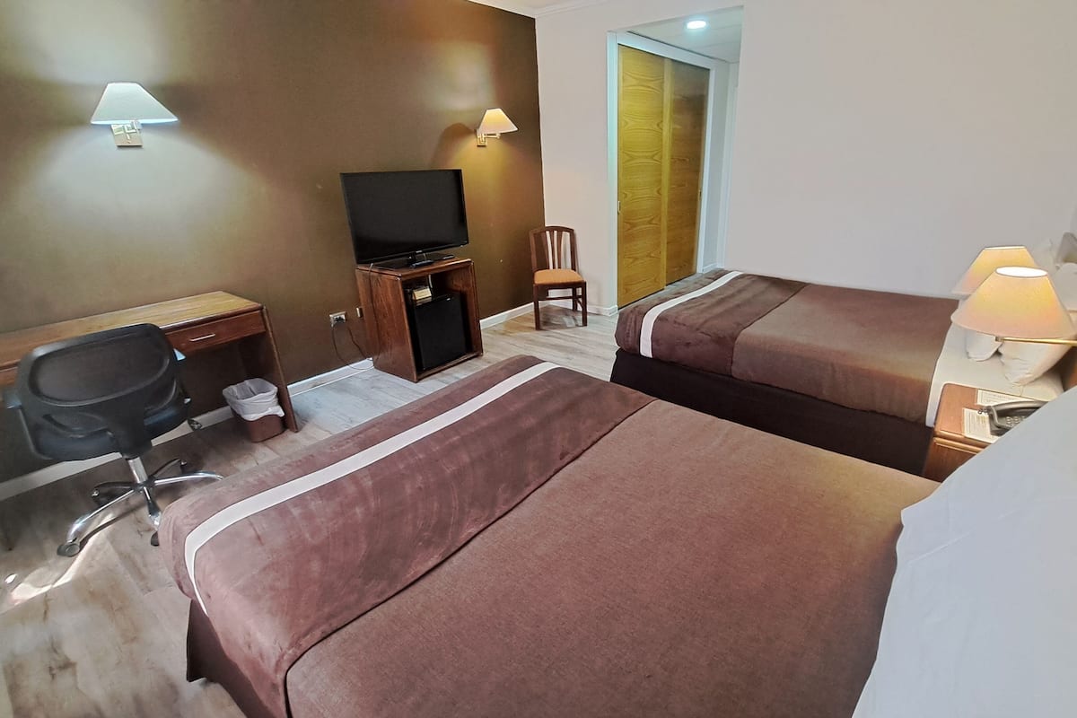 Quarto de hotel com duas camas, uma escrivaninha com cadeira, televisão e um pequeno armário.