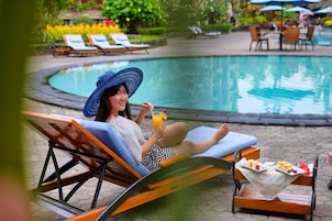 Uma pessoa relaxando em uma espreguiçadeira junto à piscina, com uma mesa de comida e bebidas por perto.