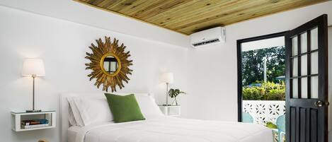 Quarto com cama, duas mesas de cabeceira com luminárias, um espelho, um cesto e vista para o exterior.