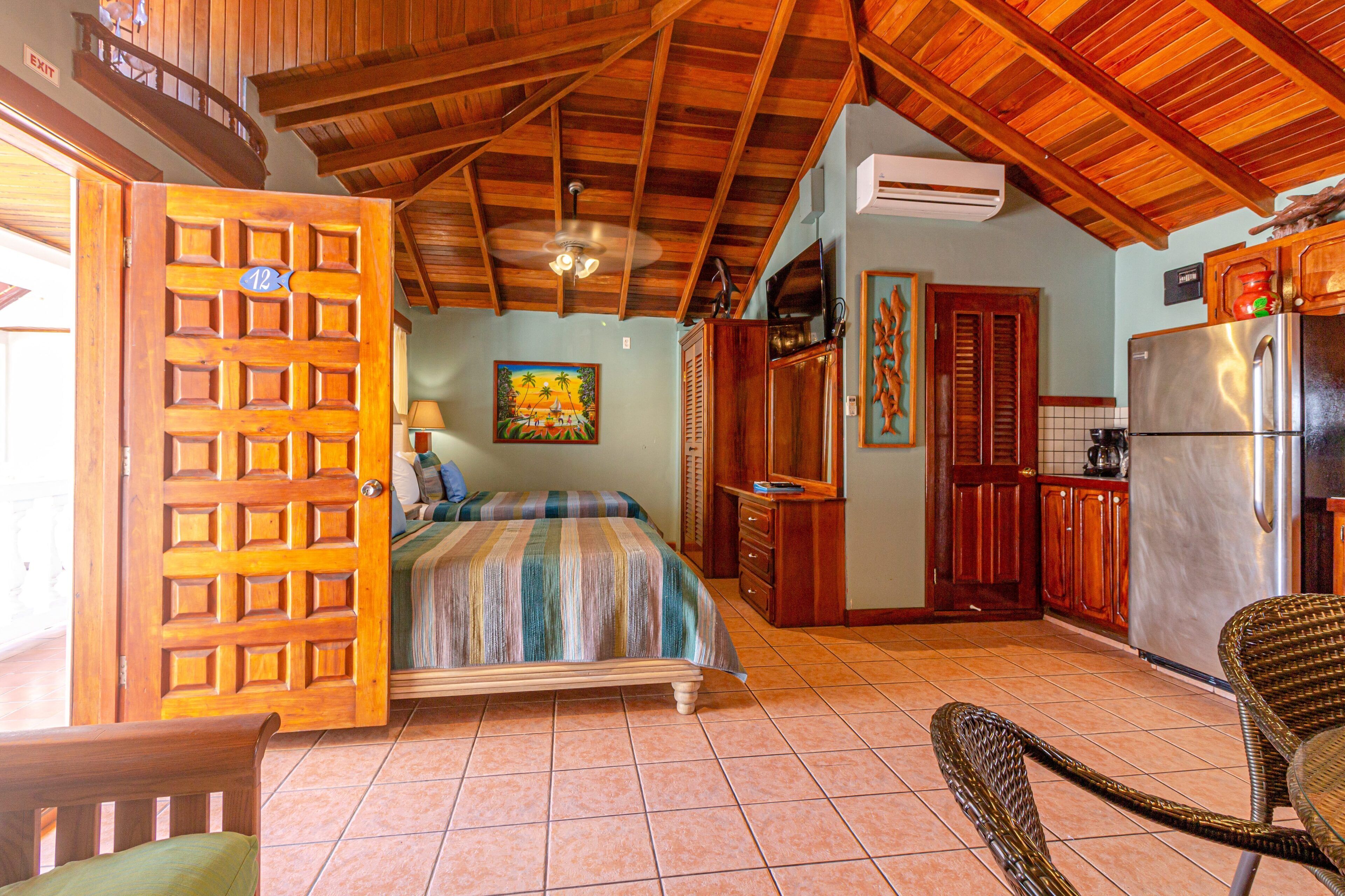 Um quarto com uma cama, porta de madeira, ventilador de teto e uma área de cozinha com geladeira.