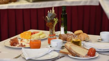 Mesa de café da manhã com pratos de comida, um vaso de flores secas, uma garrafa de azeite e um copo de suco de laranja.