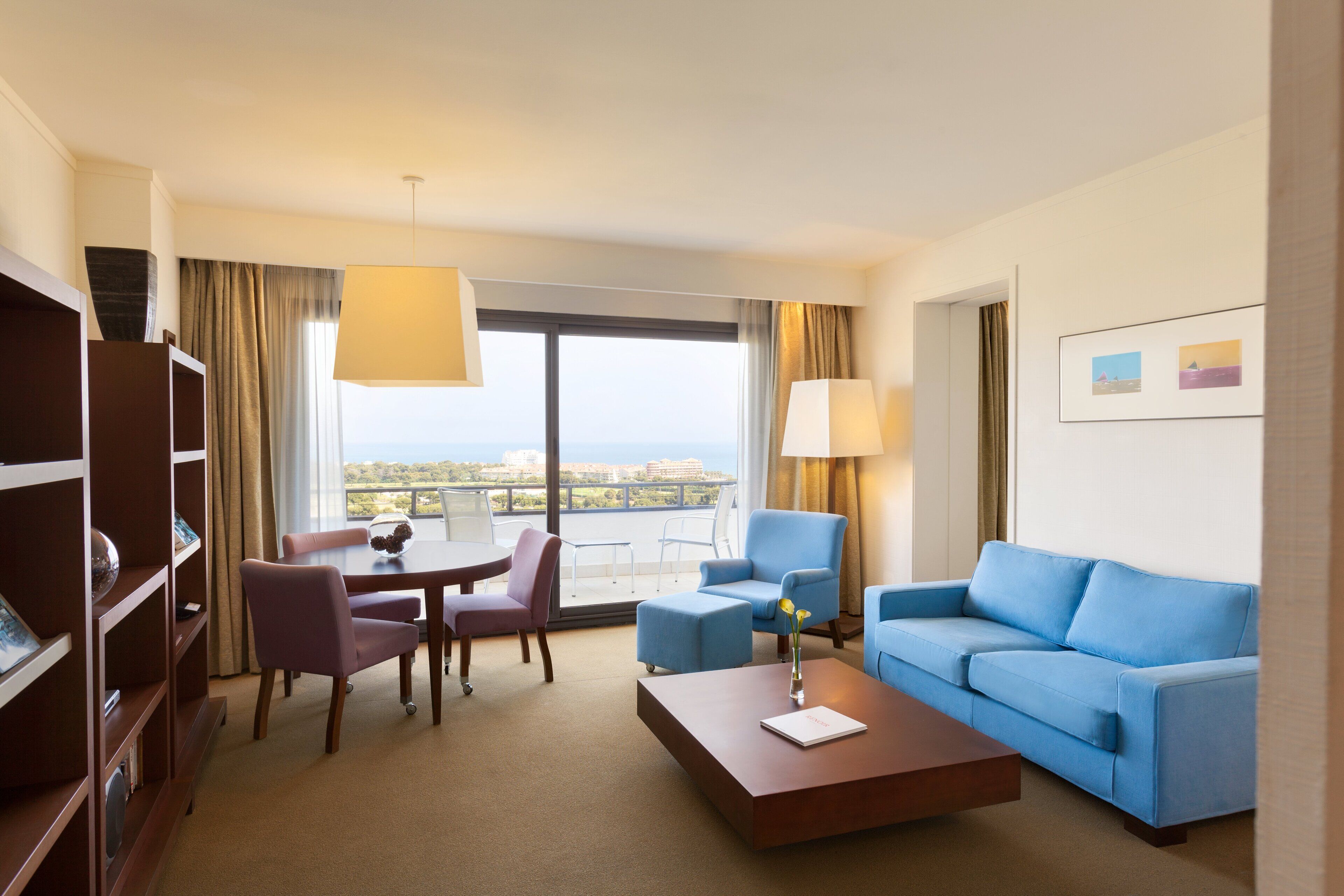 Quarto de hotel com sofá azul, mesa de centro de madeira, escrivaninha com cadeiras, estante de livros e uma grande janela com vista para a cidade.