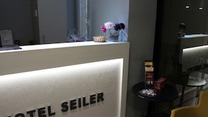 Balcão de recepção de hotel com uma placa indicando "HOTEL SEILER". Há uma pequena mesa com um cesto e um vaso com flores no balcão da recepção.