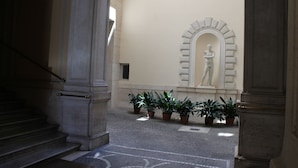 Corredor com uma estátua, plantas em vasos e arquitetura clássica.