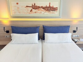 Uma cama de casal com lençóis brancos, duas almofadas azuis e um quadro emoldurado acima.