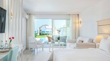 Quarto de hotel com uma cama, duas poltronas brancas, uma mesa pequena, televisão e vista para o mar através de grandes janelas.