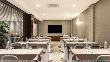 Sala de conferência com mesas preparadas para uma reunião, equipada com garrafas de água e cadernos, e uma tela grande na frente.