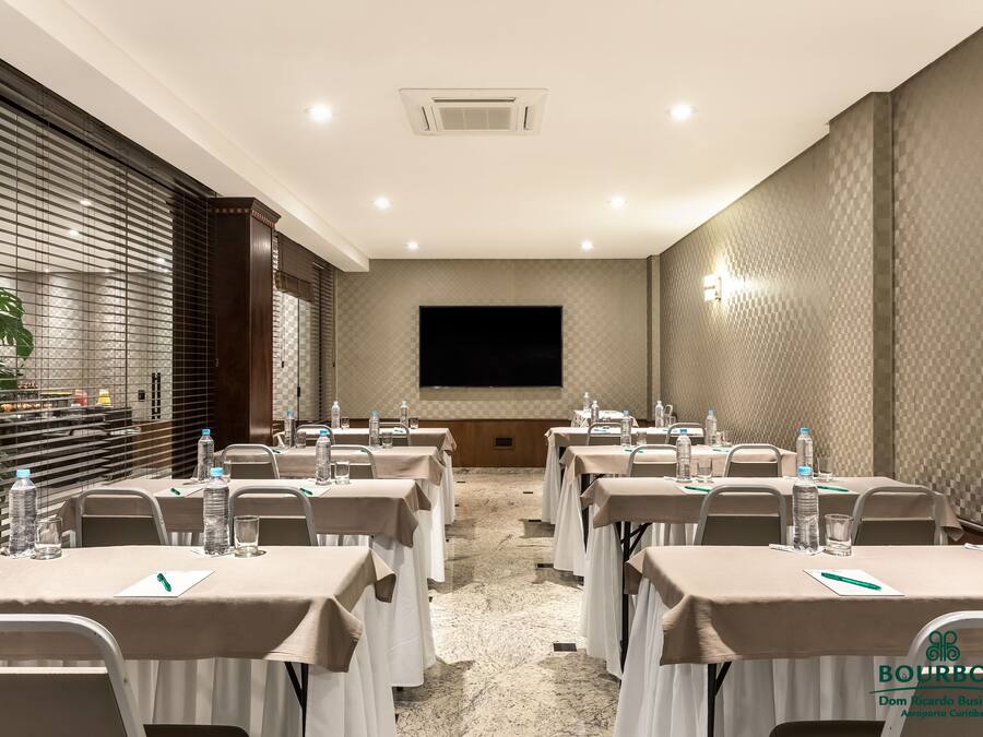 Sala de conferência com mesas preparadas para uma reunião, equipada com garrafas de água e cadernos, e uma tela grande na frente.
