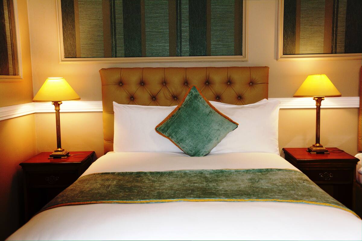 Cama bem arrumada com cabeceira estofada, duas luminárias laterais e um cobertor verde.