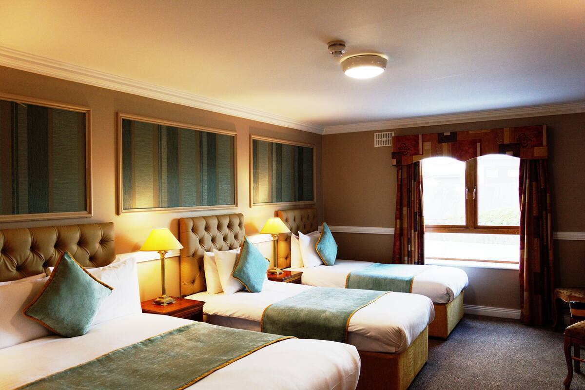 Quarto de hotel com duas camas, cabeceira, janela com cortinas e abajur no criado-mudo.