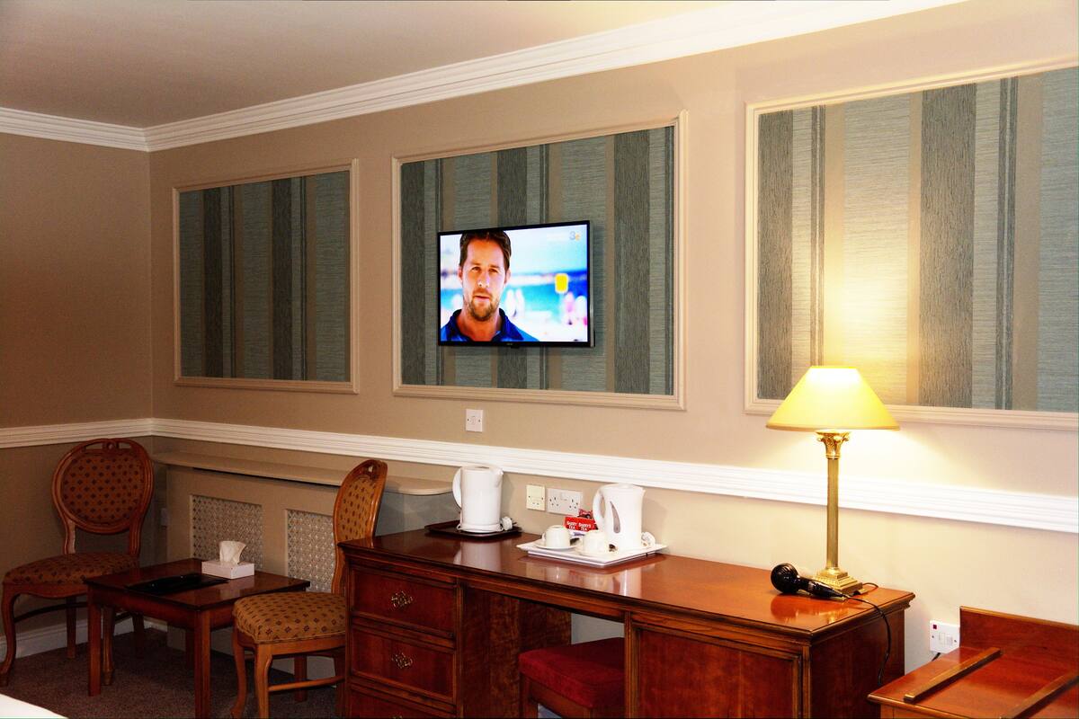 Quarto de hotel com televisão na parede, uma escrivaninha com candeeiro e duas cadeiras.