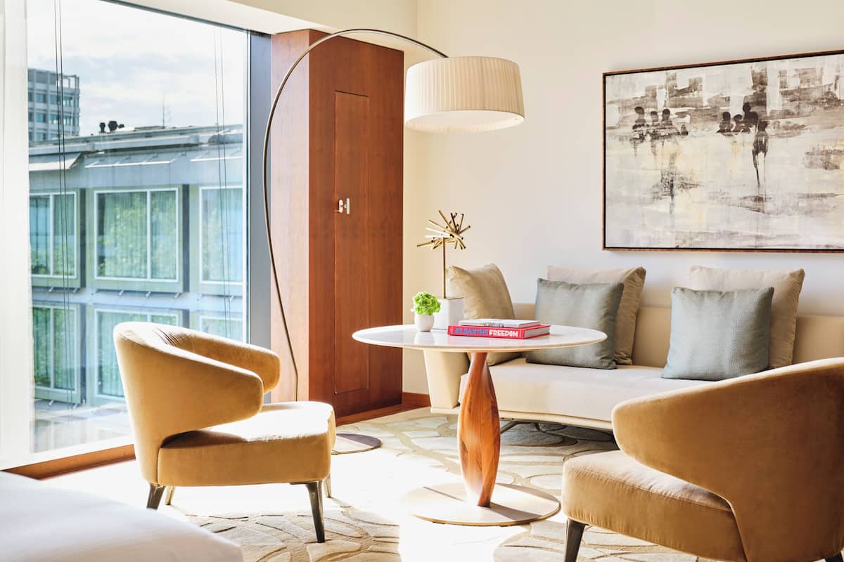 Uma sala de estar moderna com sofá, duas poltronas, uma mesa redonda com abajur, um quadro na parede e vista para edifícios através de grandes janelas.