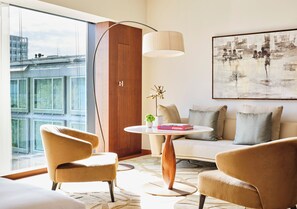 Uma sala de estar moderna com sofá, duas poltronas, uma mesa redonda com abajur, um quadro na parede e vista para edifícios através de grandes janelas.