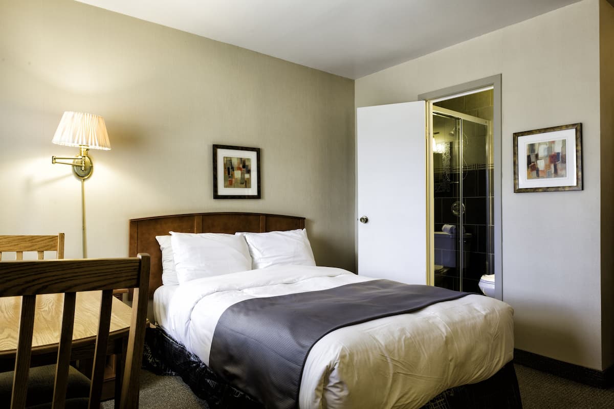 Quarto de hotel com cama, uma cadeira de madeira, escrivaninha, luminária e porta para o banheiro.