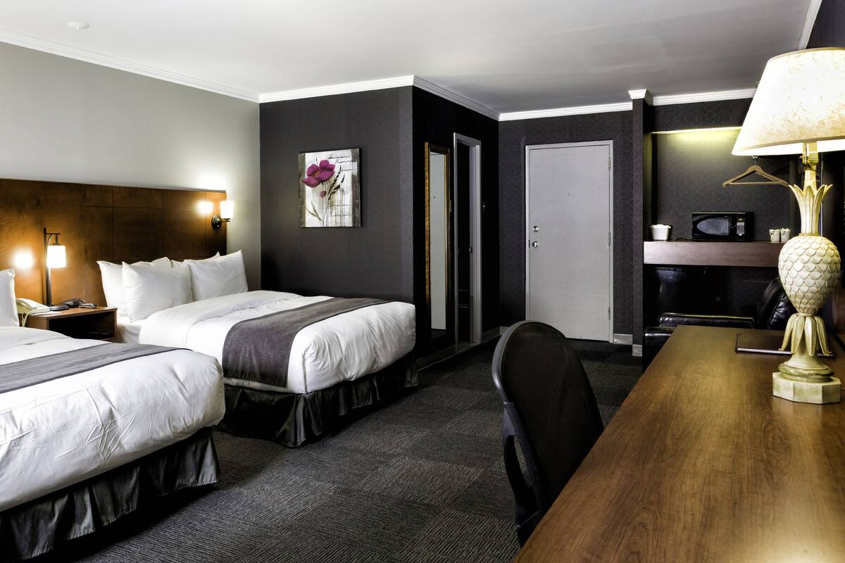 Quarto de hotel com duas camas, uma escrivaninha, uma cadeira, um abajur, um micro-ondas e porta de acesso a um banheiro.