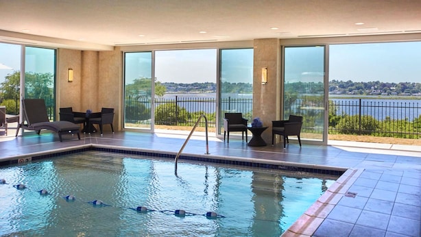 Piscina interna com água cristalina, cercada por grandes janelas que oferecem vista para um corpo d'água e vegetação.