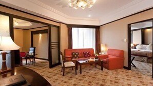 Quarto de hotel com sofá, poltrona e uma pequena mesa. Há um candelabro no teto e uma cama com cabeceira.