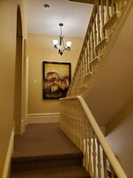 Escada revestida com carpete, com corrimão branco, um candelabro e um quadro emoldurado na parede.