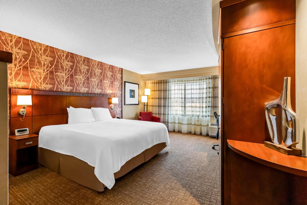 Quarto de hotel com uma cama grande, cabeceira de madeira, mesa de cabeceira com abajur, uma cadeira vermelha e uma escrivaninha com espelho.