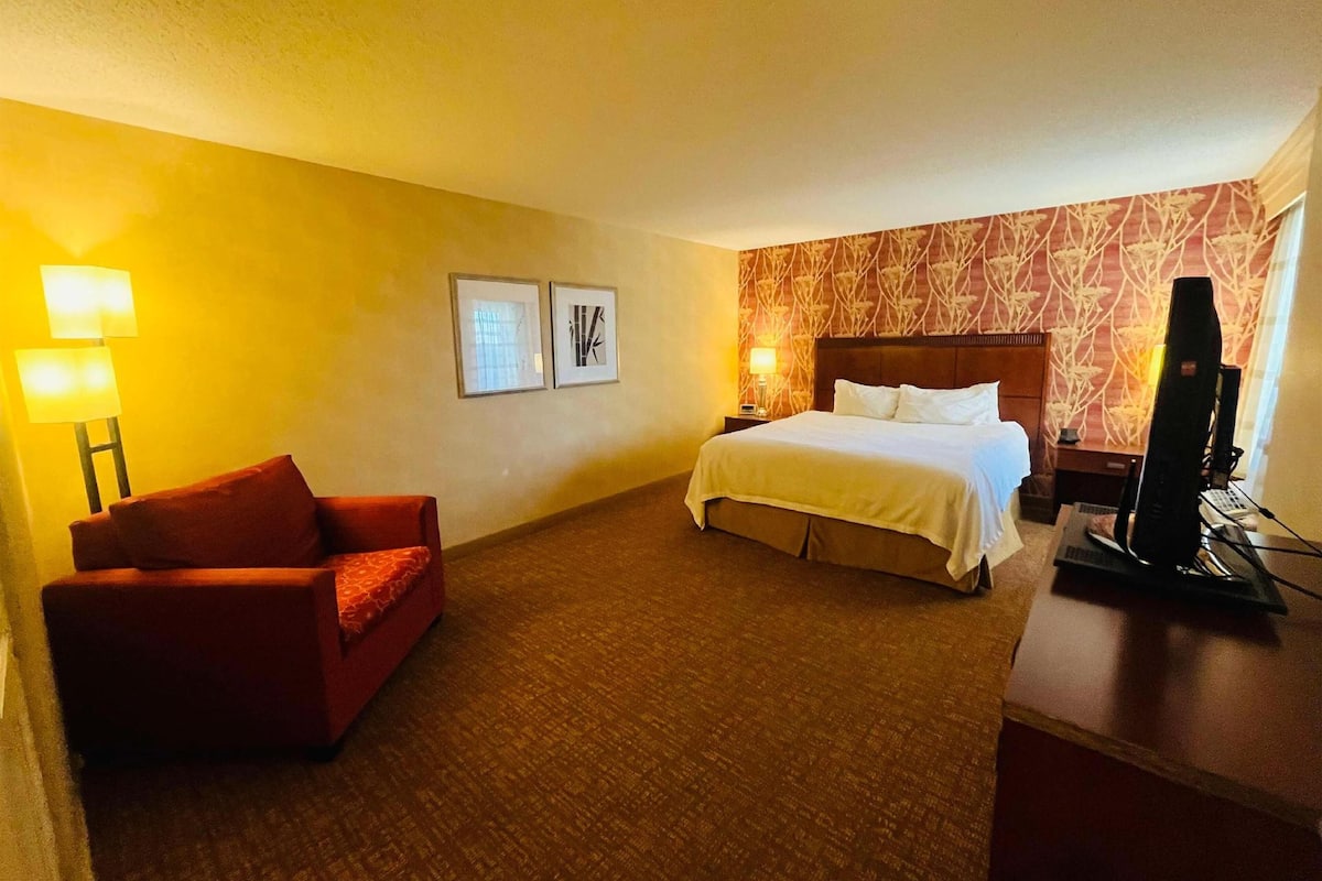 Quarto de hotel com uma cama, uma poltrona vermelha, uma escrivaninha com um computador e duas fotos emolduradas na parede.