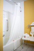 Casa de banho com banheira branca, cortina de chuveiro, barra de apoio, lavatório branco com toalha e parede amarela.